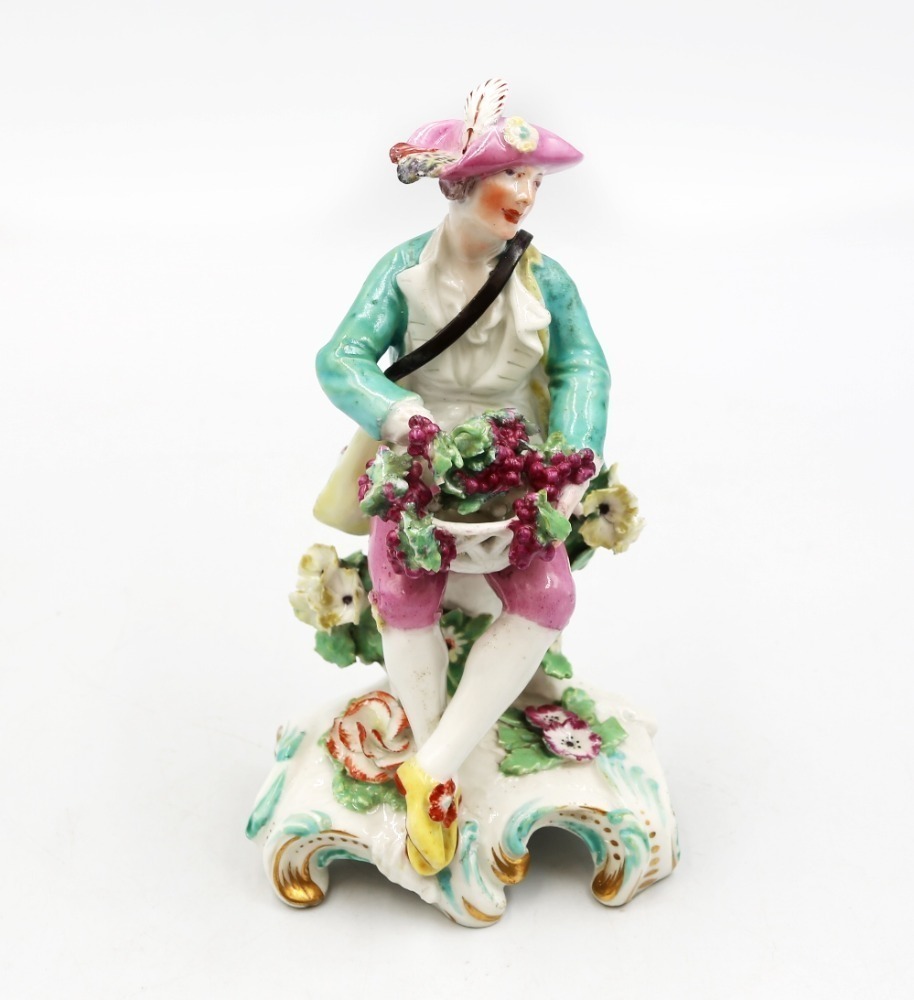 A Derby figure of a ‘grape seller’ seated on a tree stump holding a ...